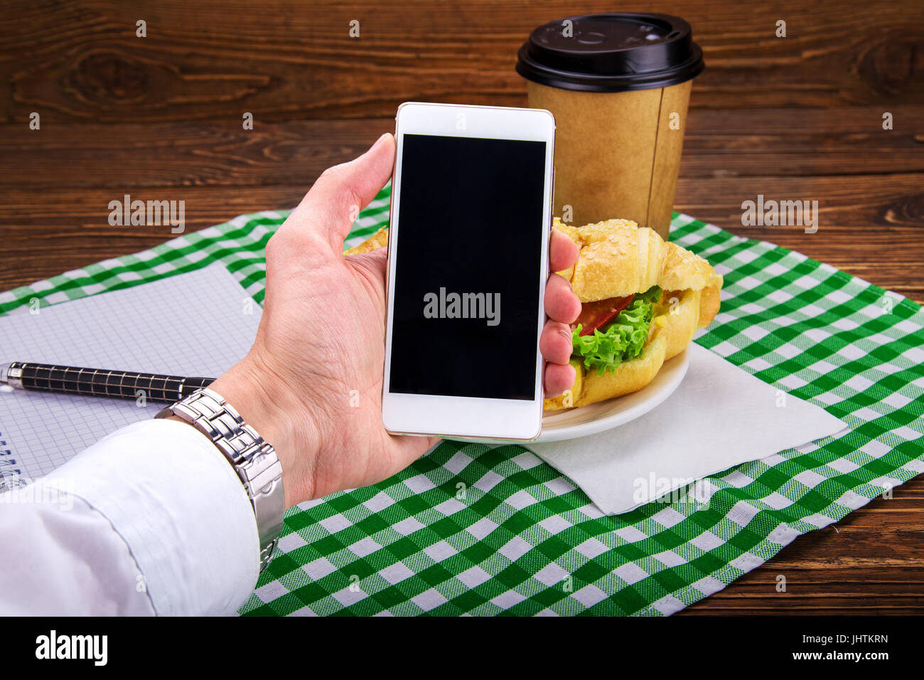 Croissant sandwich on a saucer. Mobile phone in hand. Notepad and pen ...