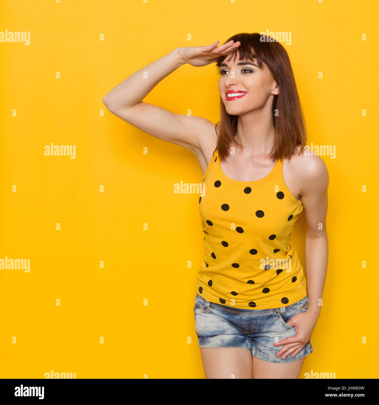 Young cheerful woman in yellow dotted shirt, jeans shorts holding hand ...