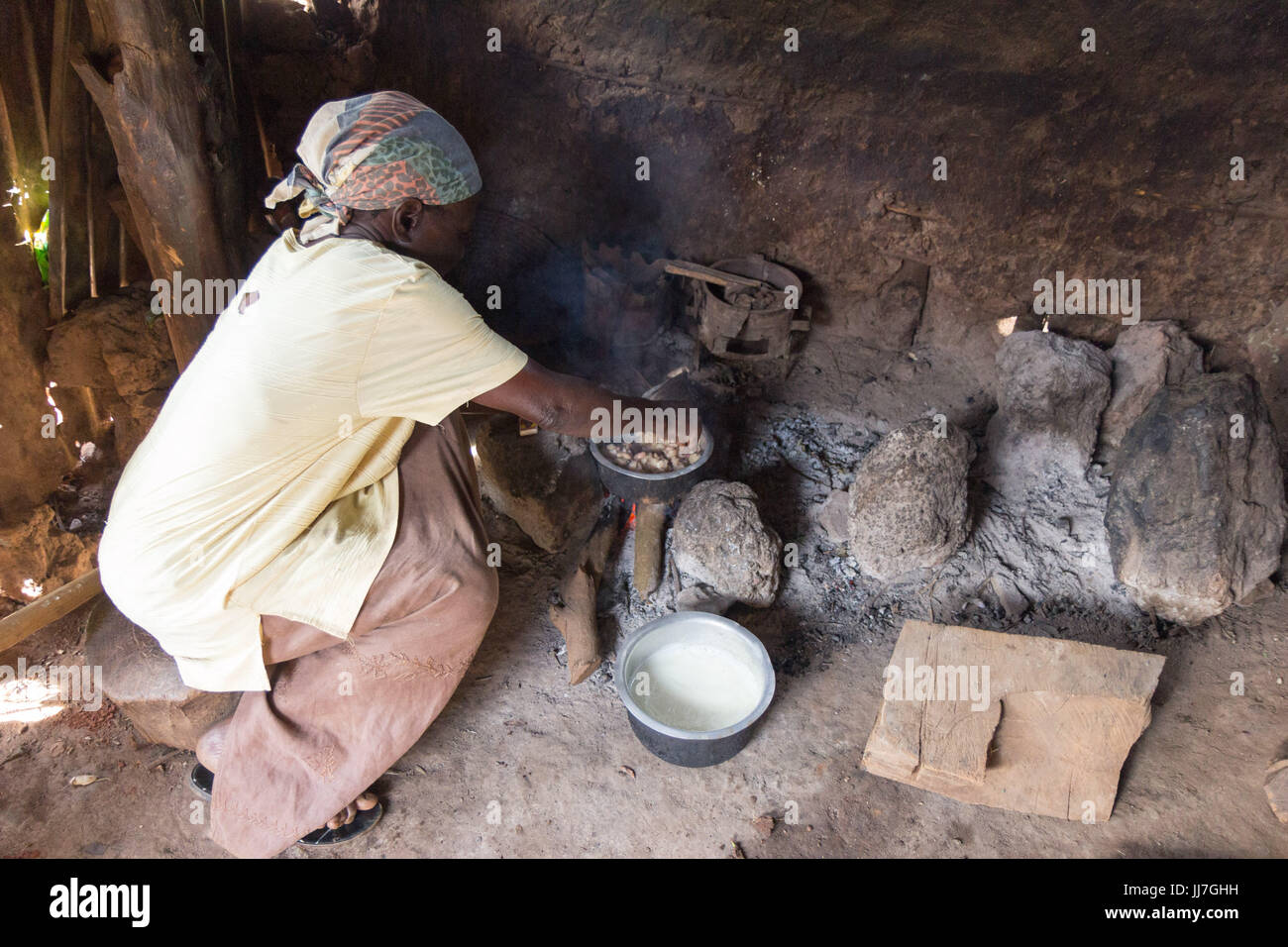 An old black Ugandan woman cooking in metal pots on charcoal fire in a ...