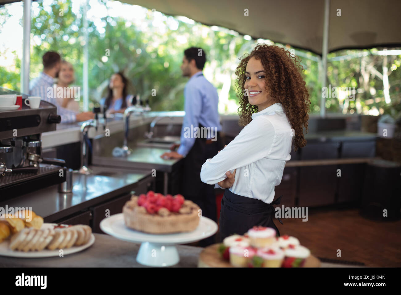 Portrait of smiling waitress standing with arms crossed at counter in ...