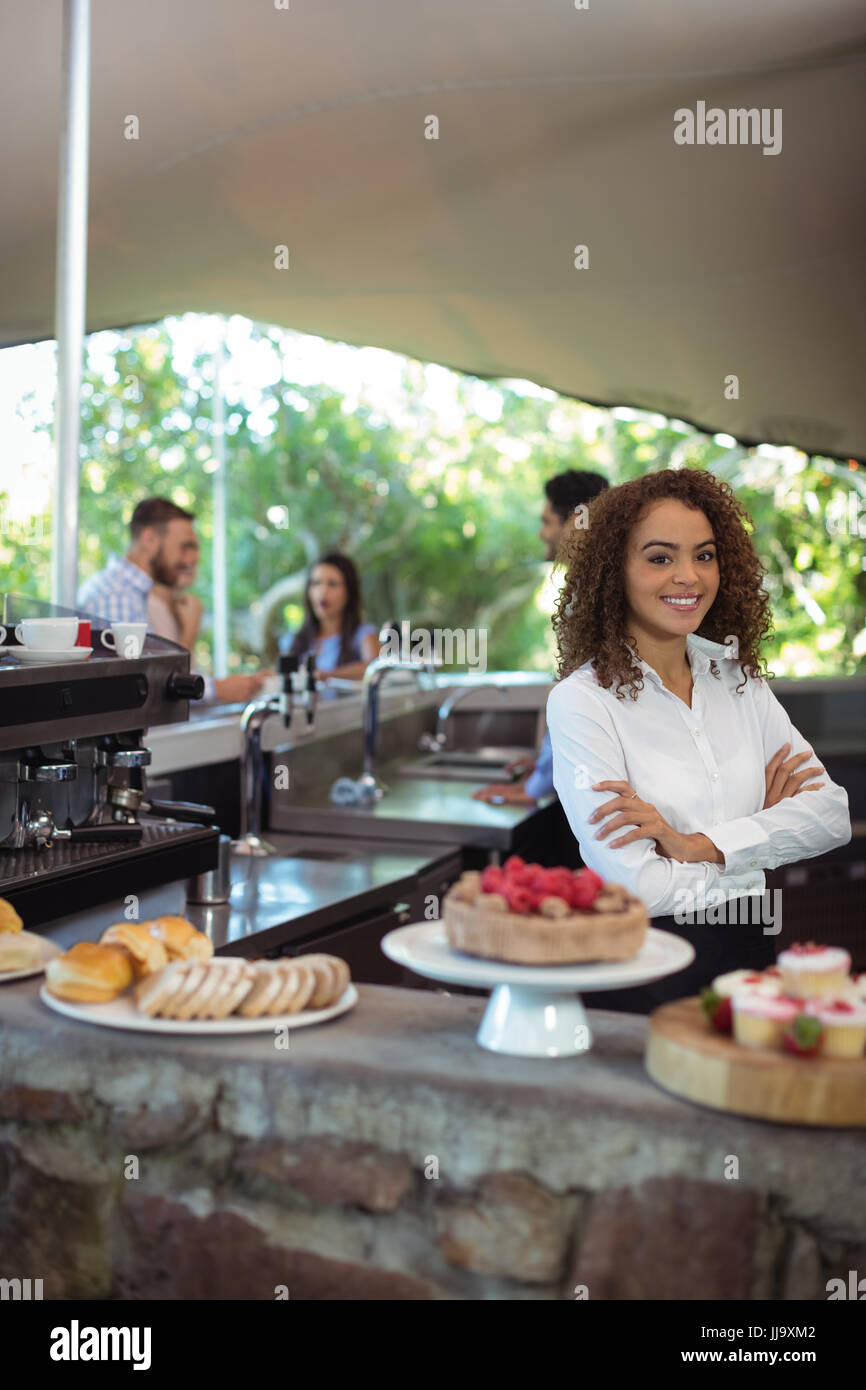 Portrait of smiling waitress standing with arms crossed at counter in ...