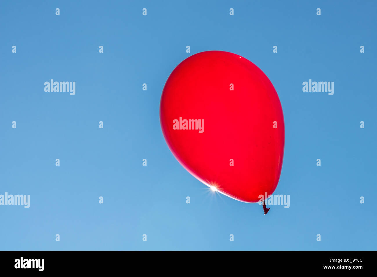 Horizontal photo of a bright red balloon against a blue sky and the sun ...