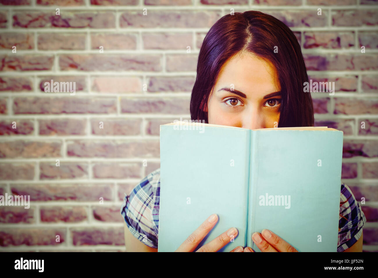 Close up portrait of woman hiding behind book against image of a wall ...
