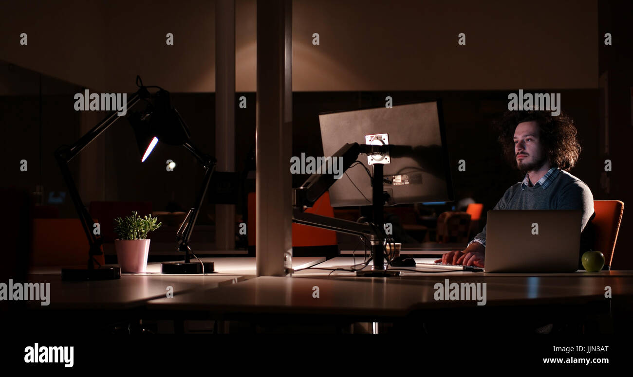 Young man working on computer at night in dark office. The designer ...