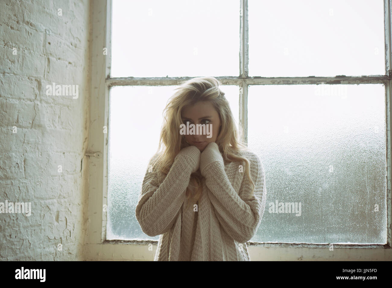 Scared young woman wearing a wool jumper standing by the window looking ...