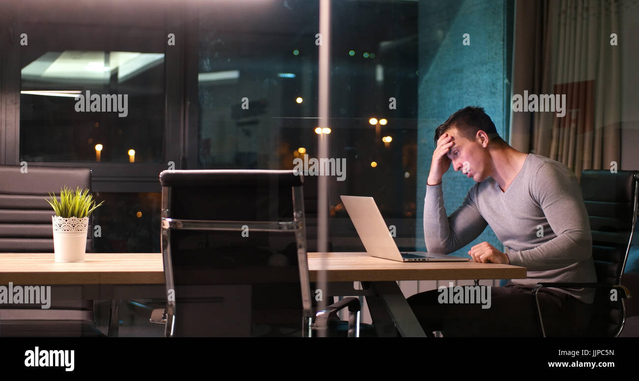 Young man working on laptop at night in dark office. The designer works ...
