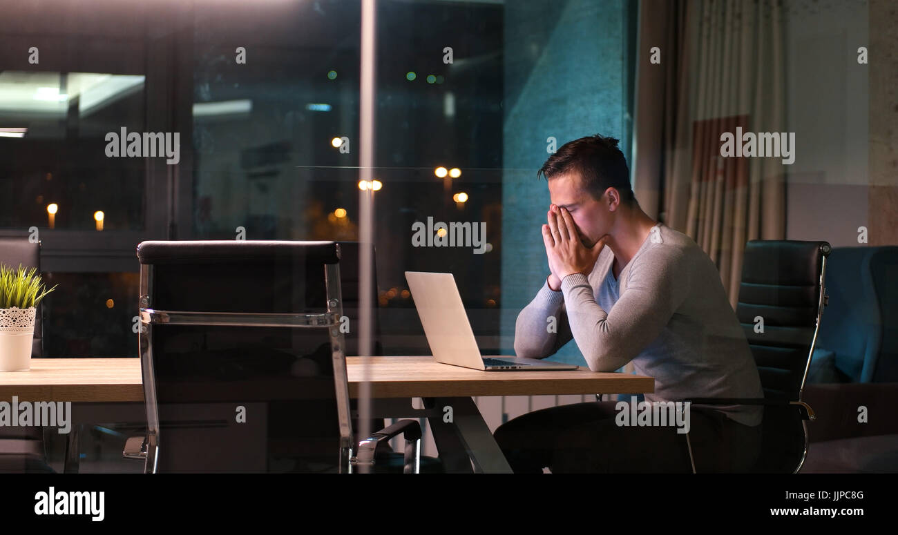 Young man working on laptop at night in dark office. The designer works ...
