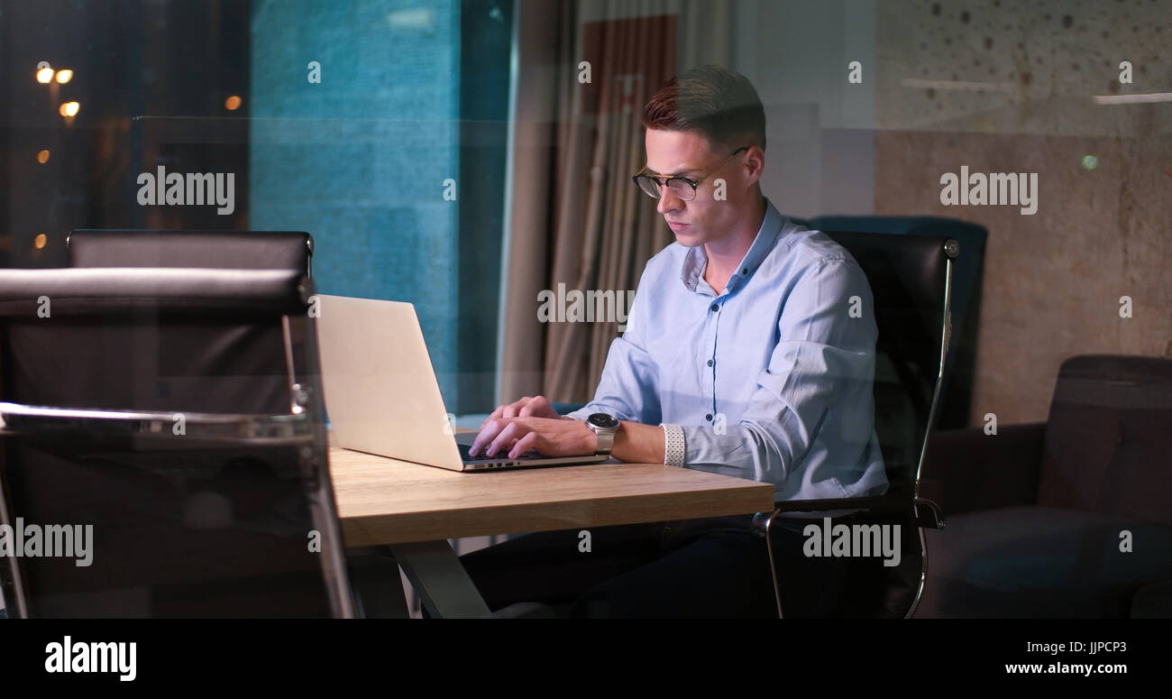 Young man working on laptop at night in dark office. The designer works ...