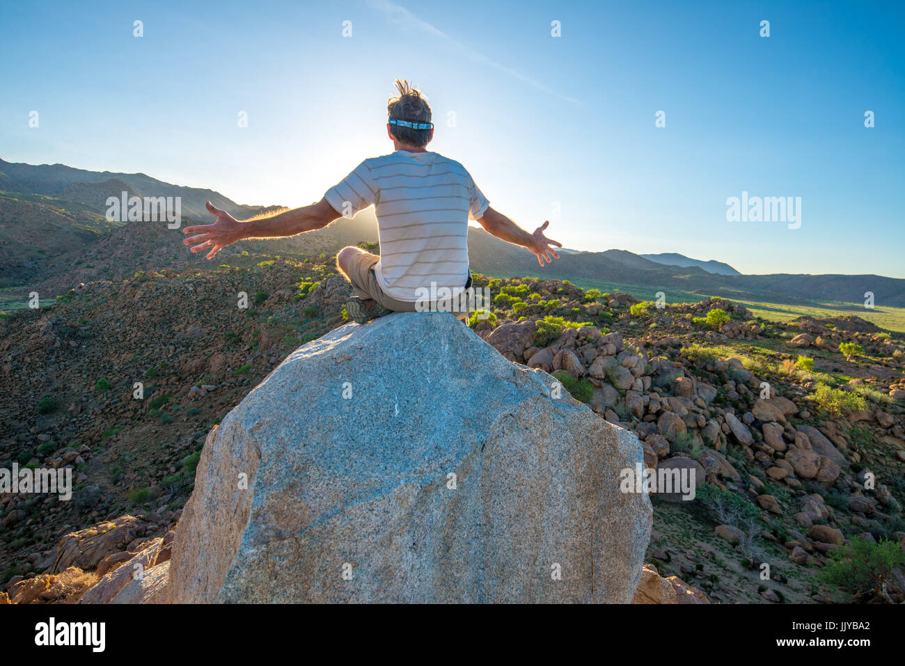A man sits on top of a large boulder with arms outstretched above the ...