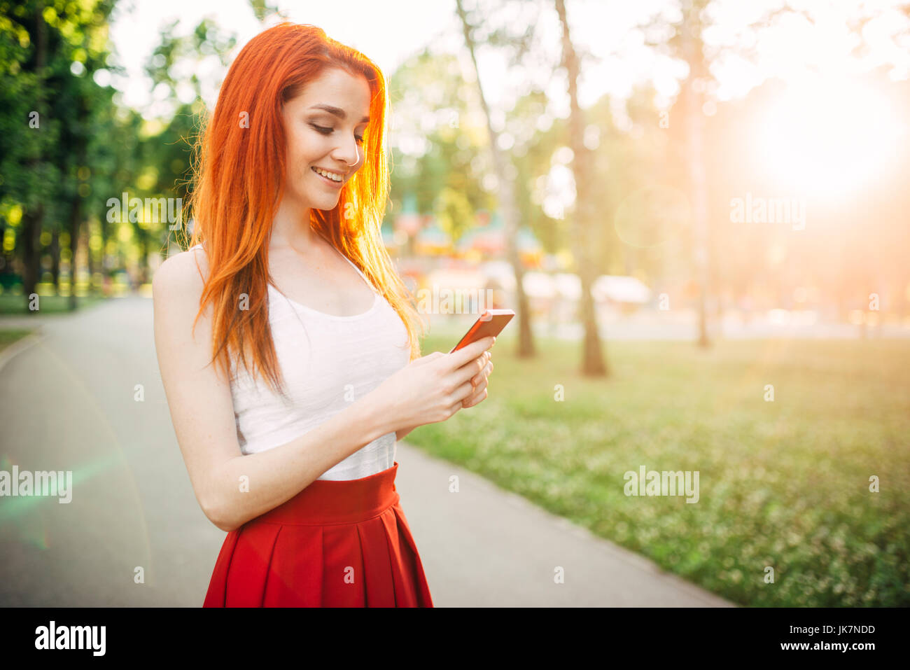 Smiling young woman with mobile phone outdoor in park on sunset. Happy ...