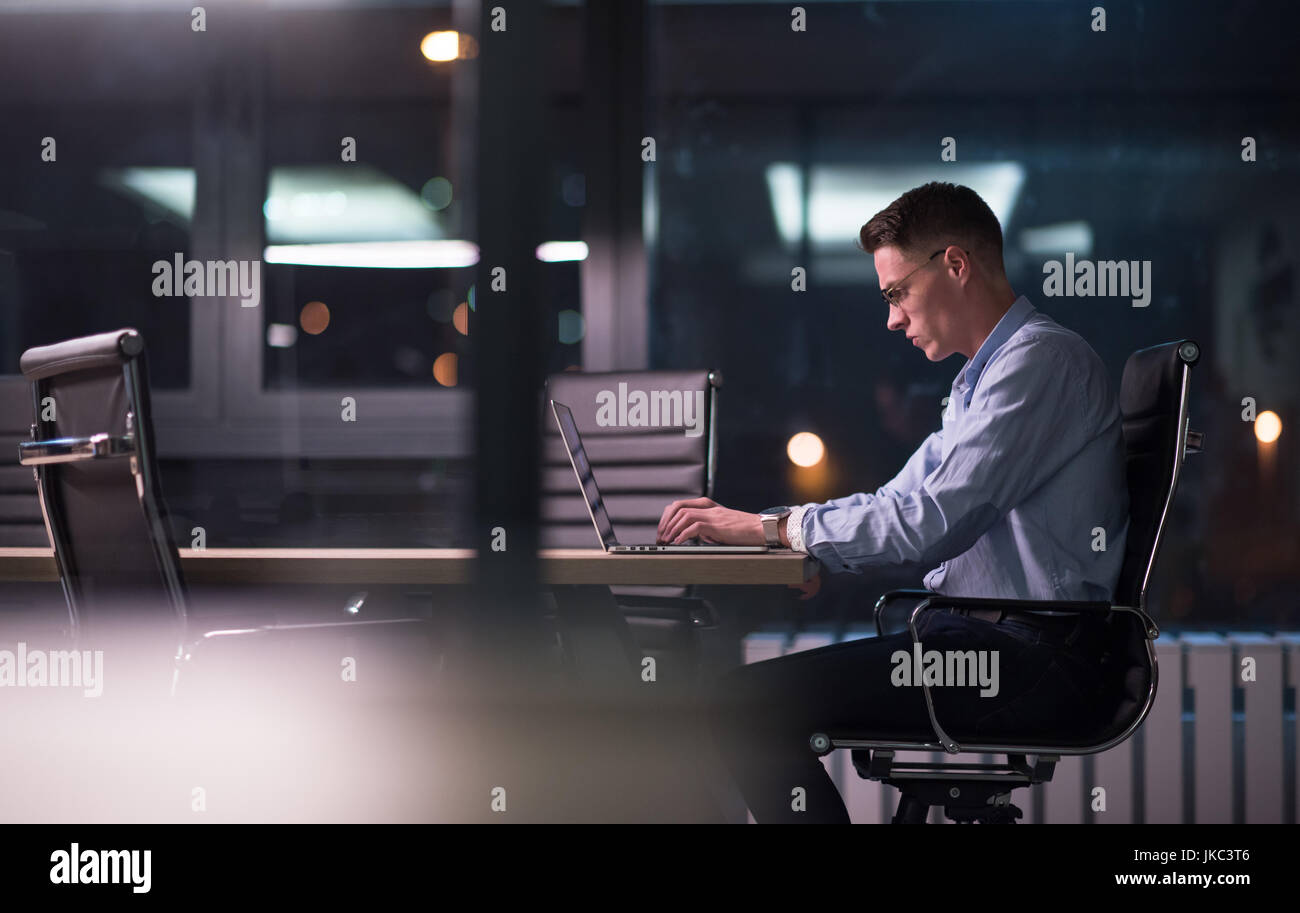 Young man working on laptop at night in dark office. The designer works ...