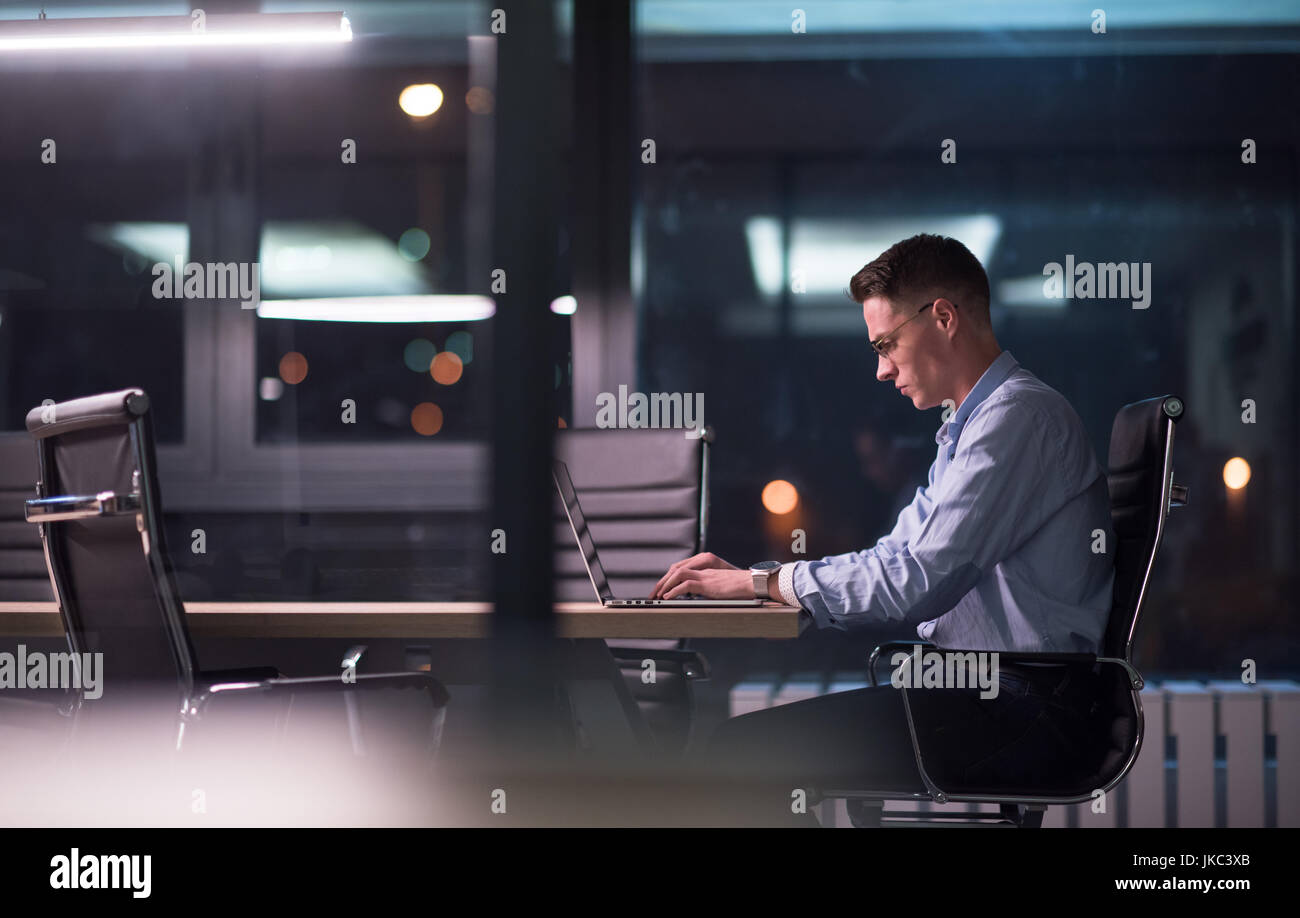 Young man working on laptop at night in dark office. The designer works ...