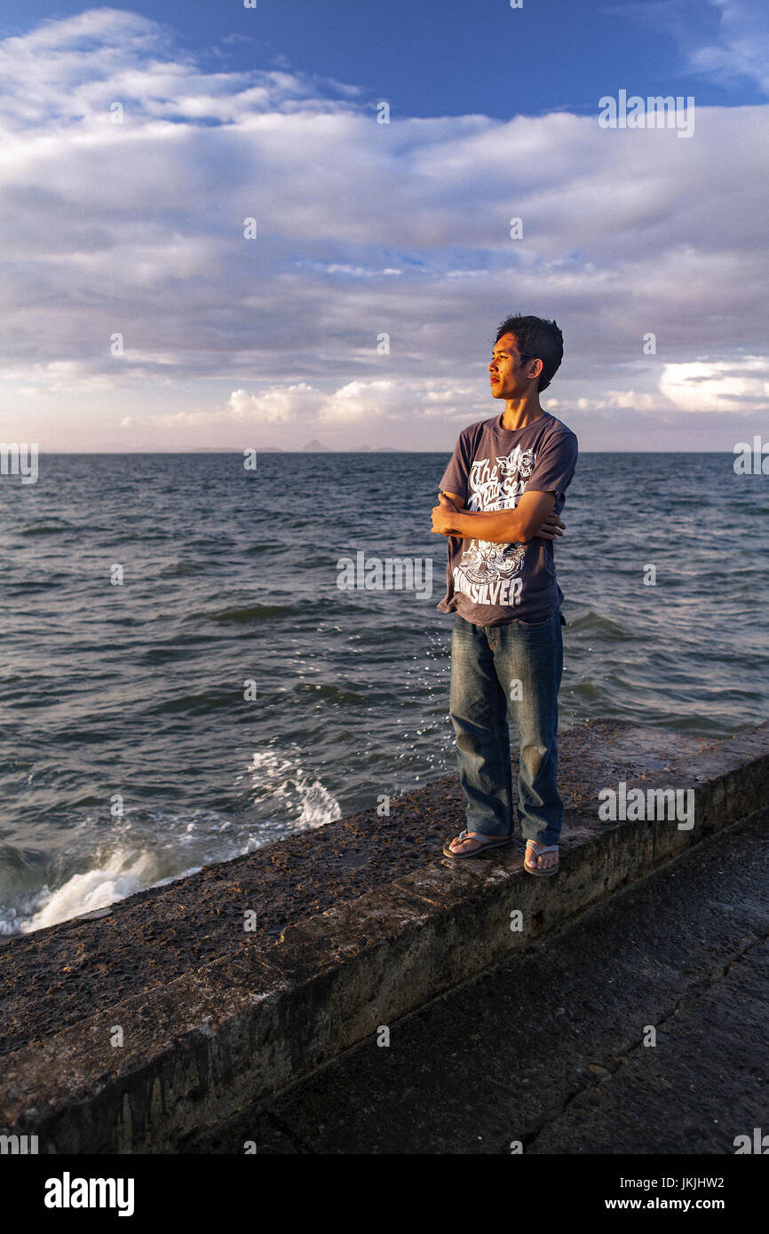 Portrait of a young Filipino man standing on a seawall watching sunset ...