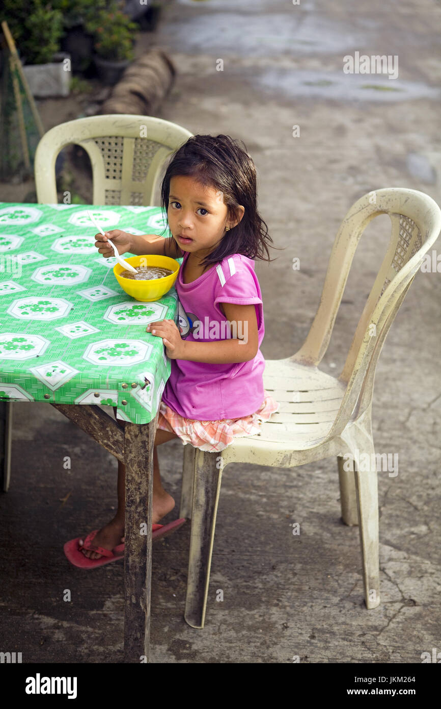 A young Filipino girl in a pink shirt sits at a table eating her ...
