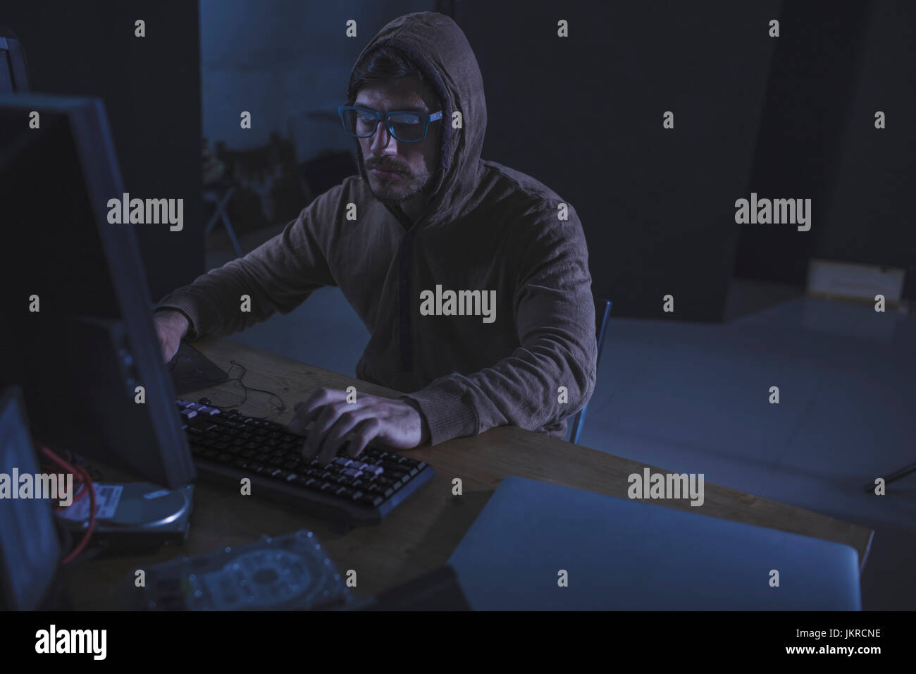 Serious computer hacker wearing hooded shirt using desktop computer at ...
