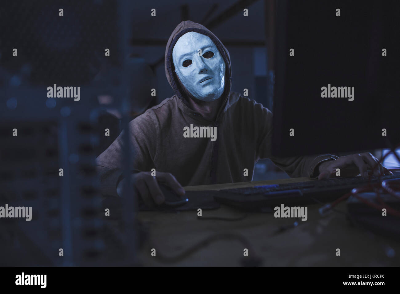 Computer hacker wearing mask and hood using computer while sitting at ...