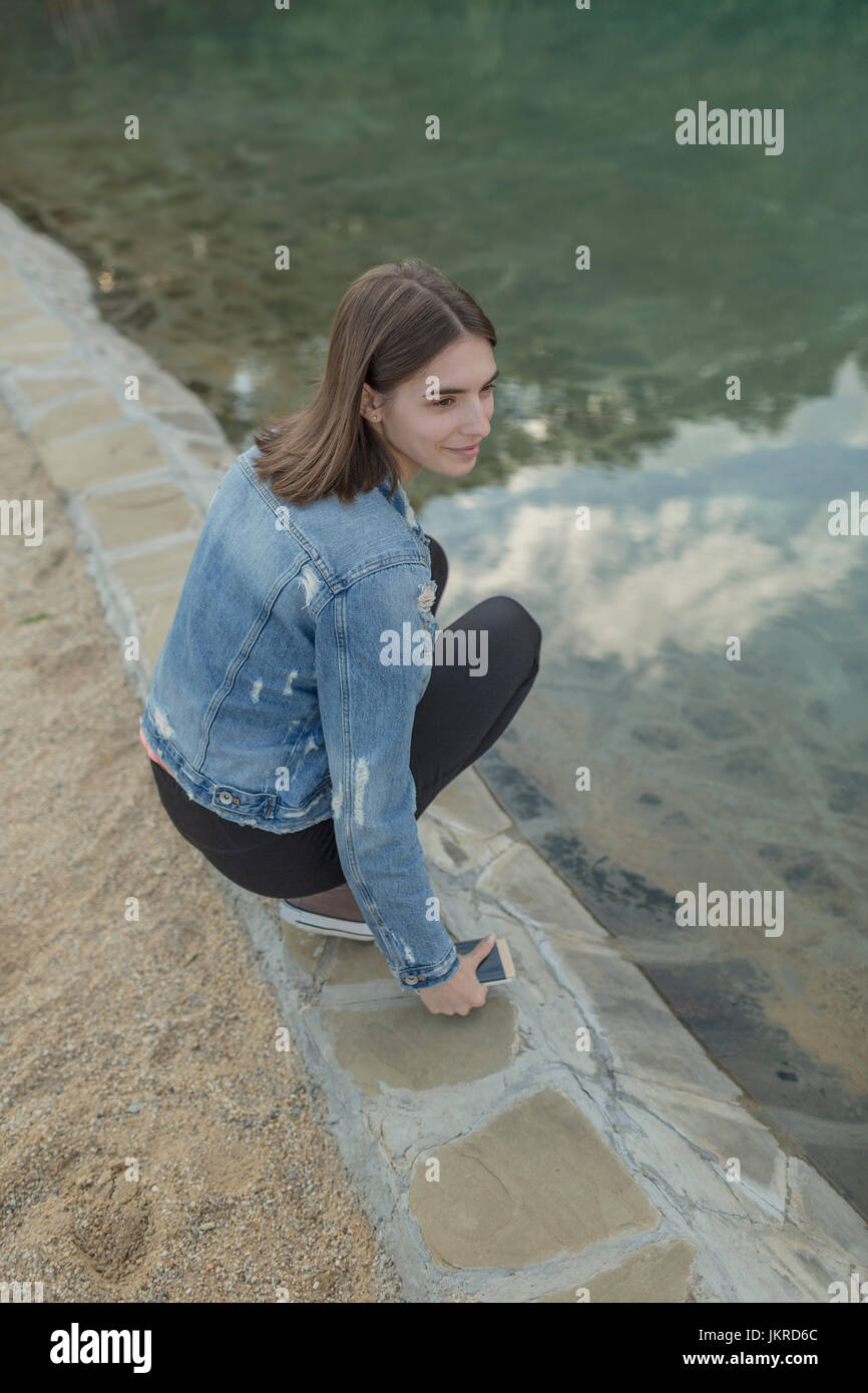 High angle view of smiling woman crouching on retaining wall by lake at ...