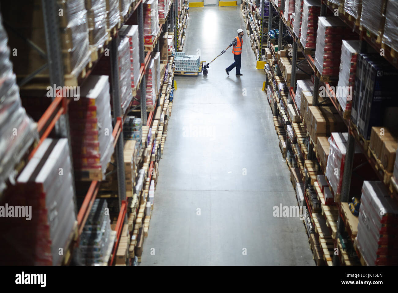 Side view of warehouse worker pulling moving cart between tall shelves ...