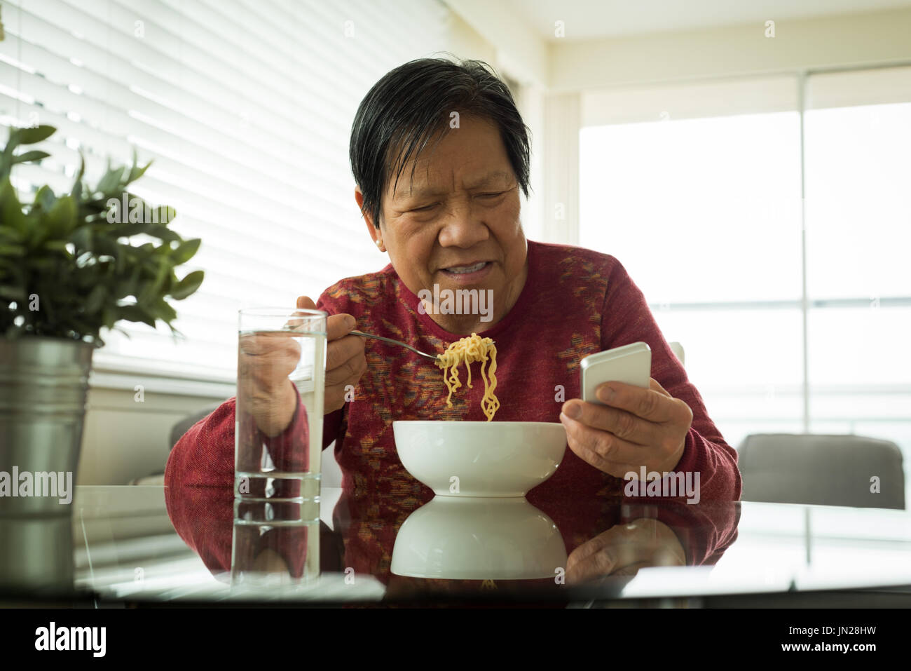 Senior woman using mobile phone while having noodles at home Stock ...
