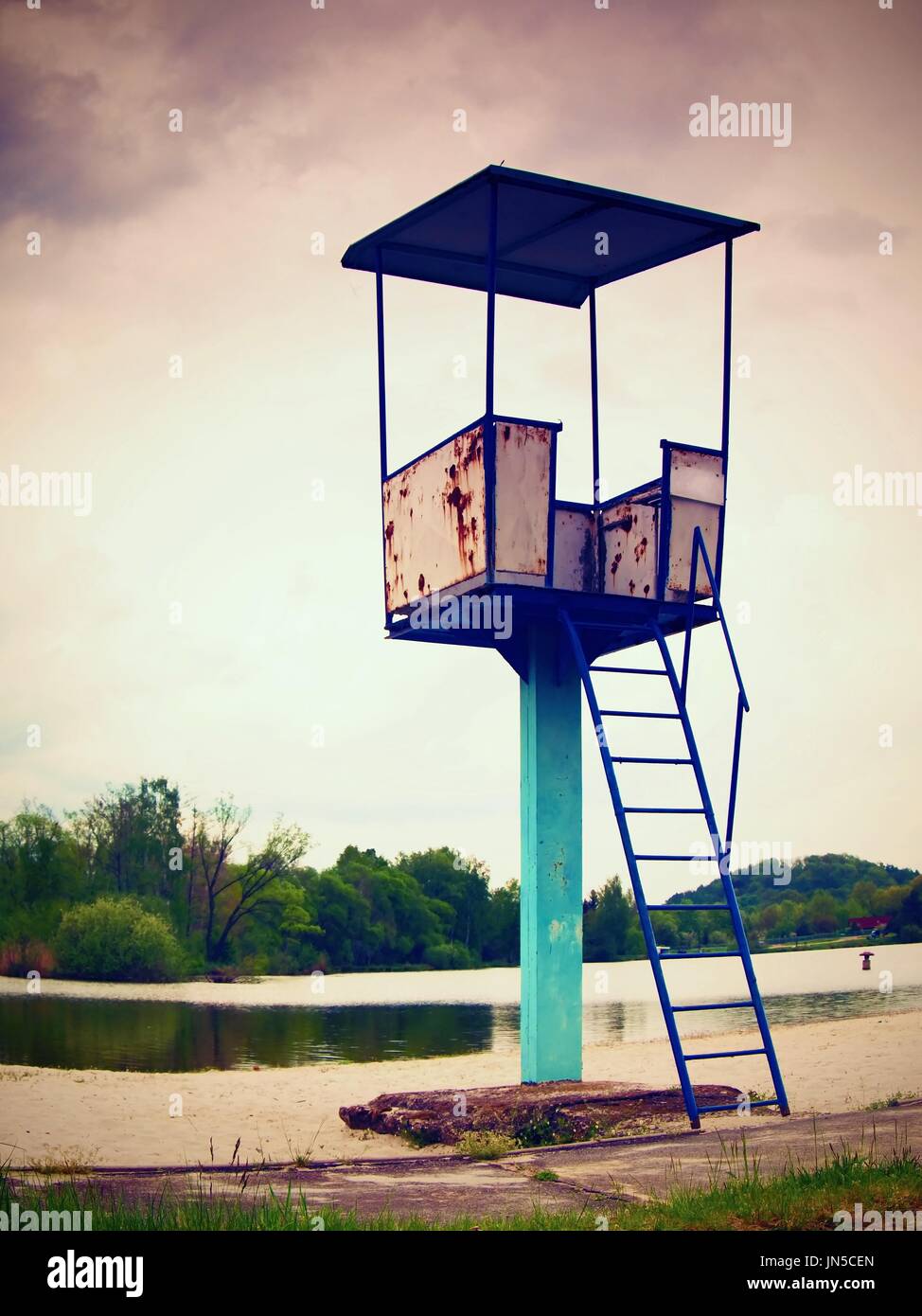 An old white and rusty metal lifeguard tower with chair on a pond beach ...