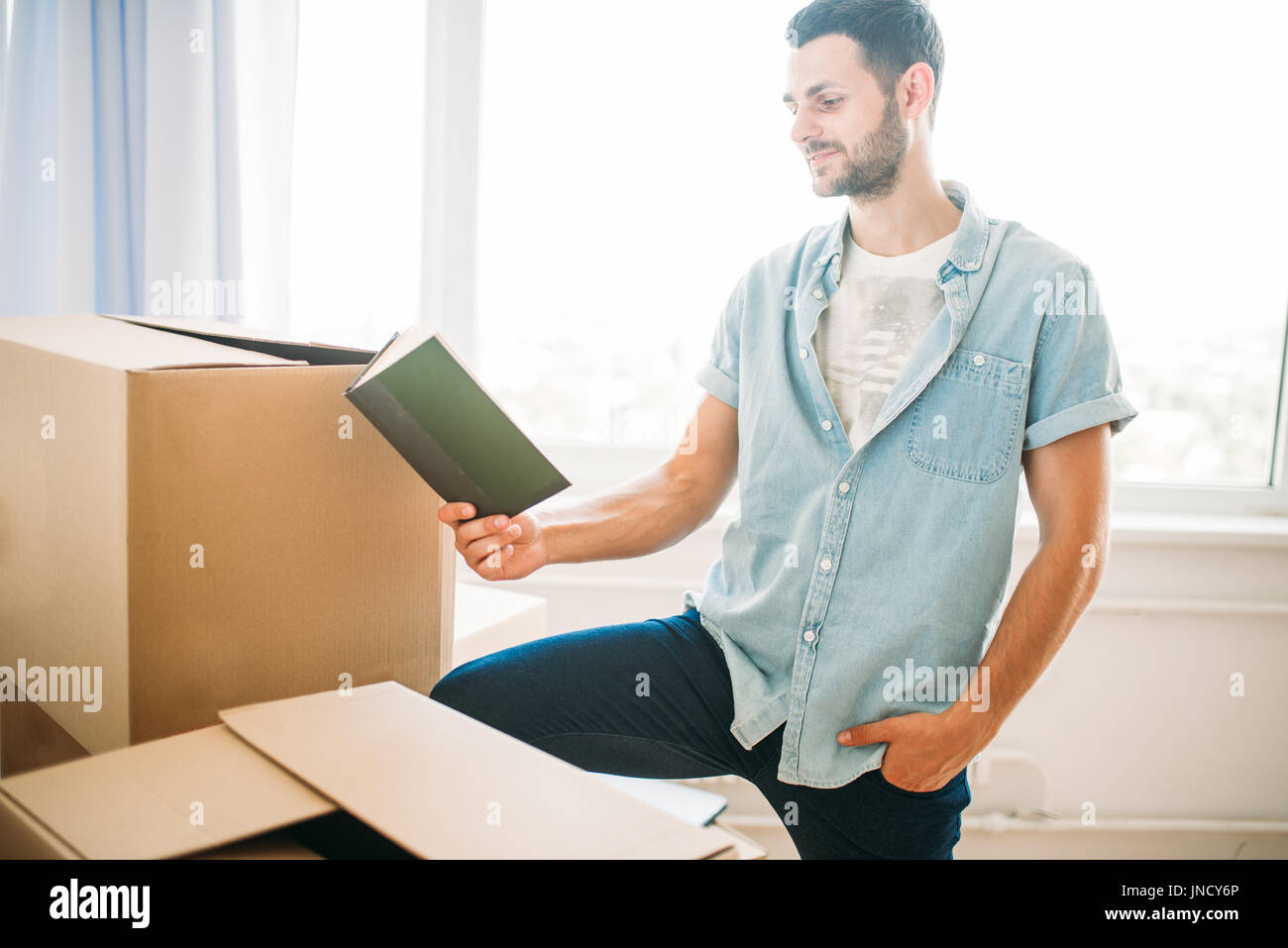 Young happy man poses with book among cardboard boxes, housewarming ...
