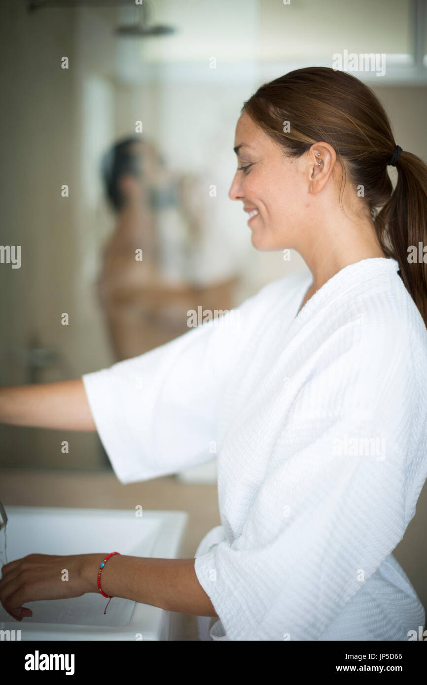 A woman standing at a sink in a bathroom, with a man taking a shower in ...