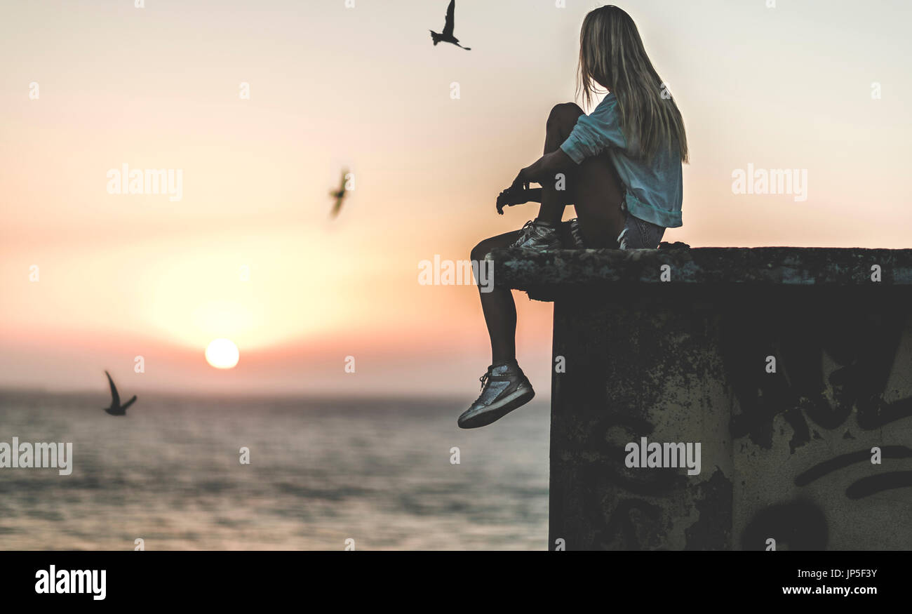 Side view of a young woman sitting on a wall with a sunset behind Stock ...
