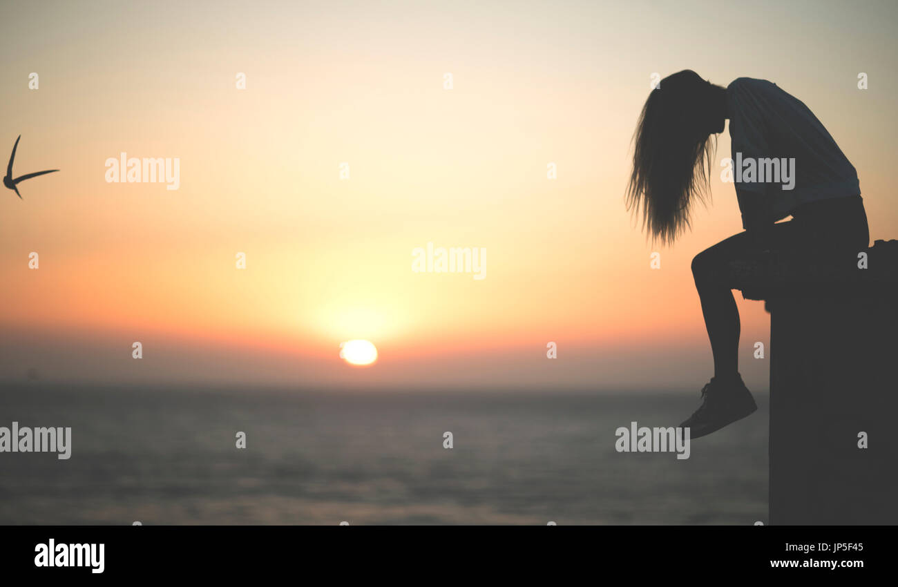 Side view of a young woman sitting on a wall with a sunset behind Stock ...