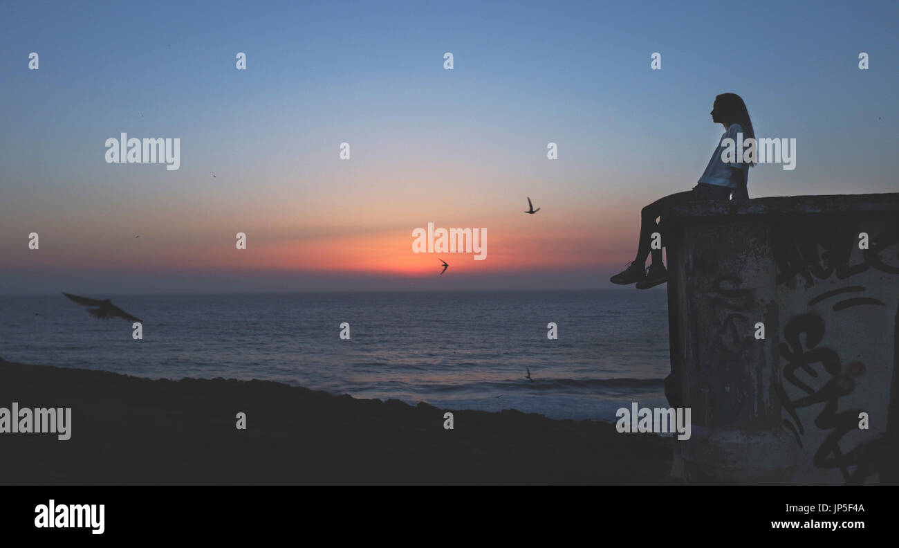 Side view of a young woman sitting on a wall with a sunset behind Stock ...