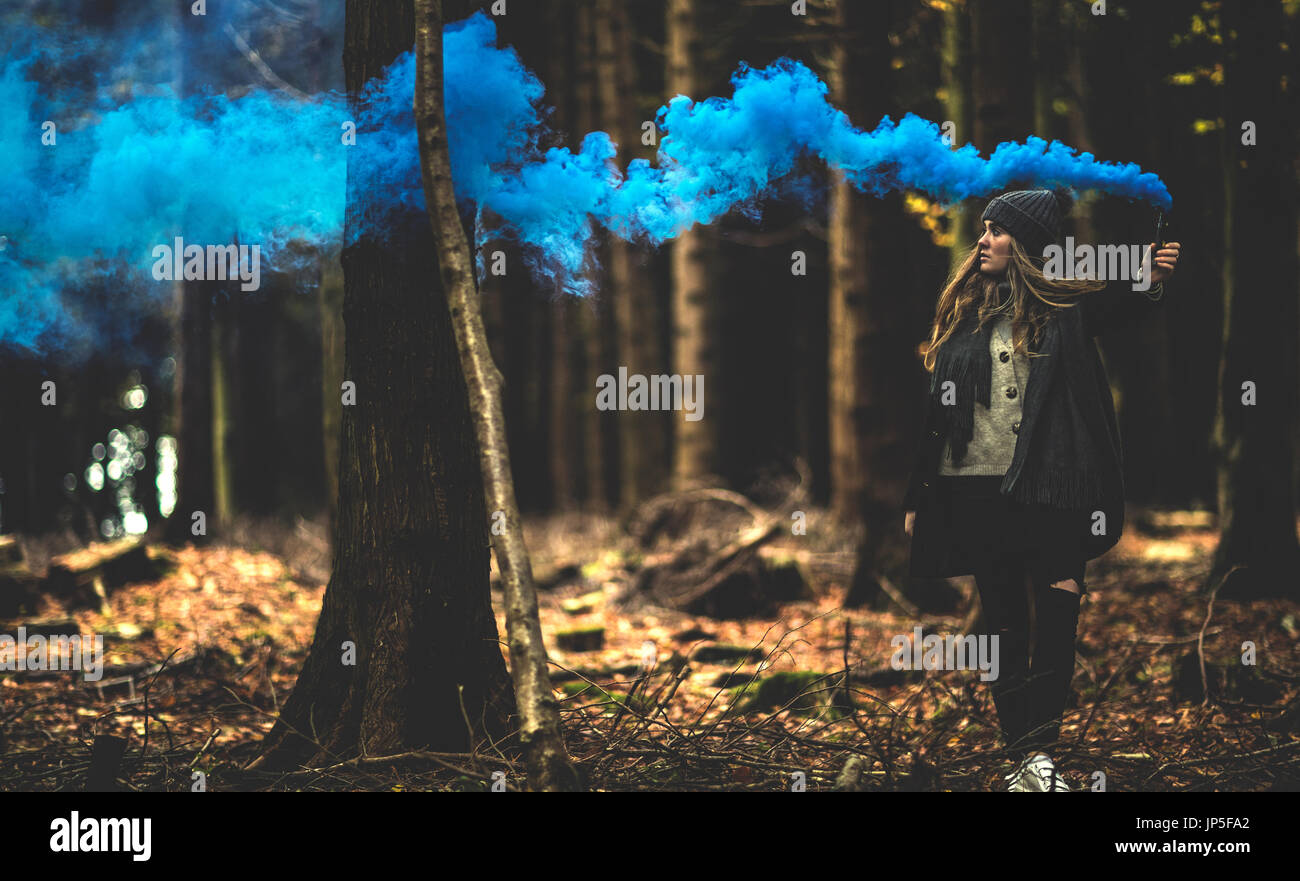 A young woman running through a forest making a blue smoke trail with a ...