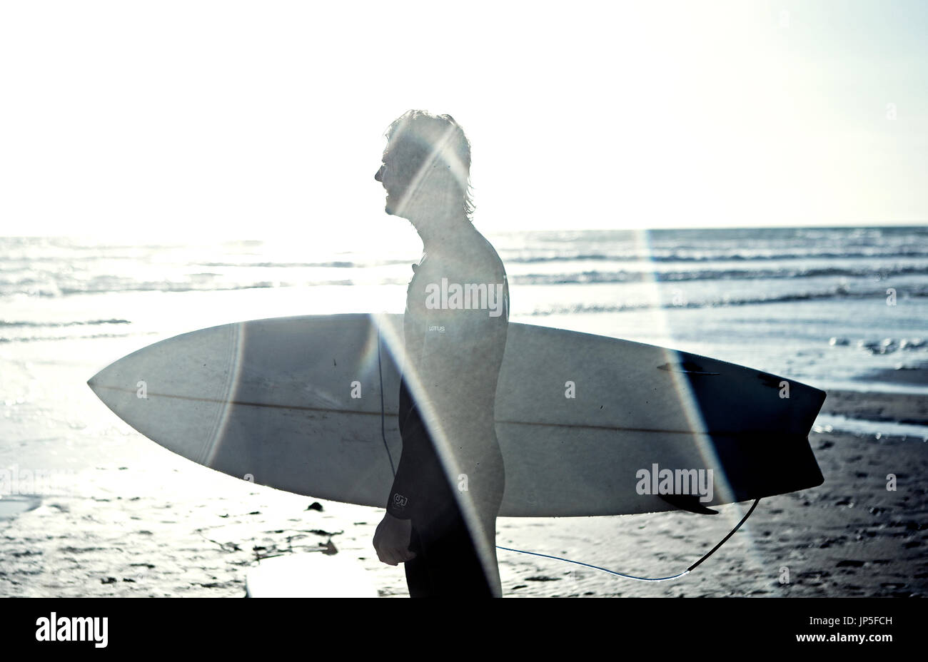 Side view of man in wetsuit standing by the ocean, carrying surfboard ...