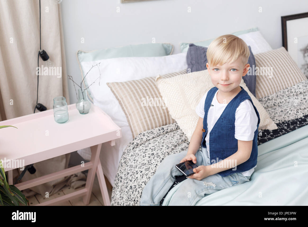 Smart small boy with mobile phone, seats on bed in bedroom. Technology ...