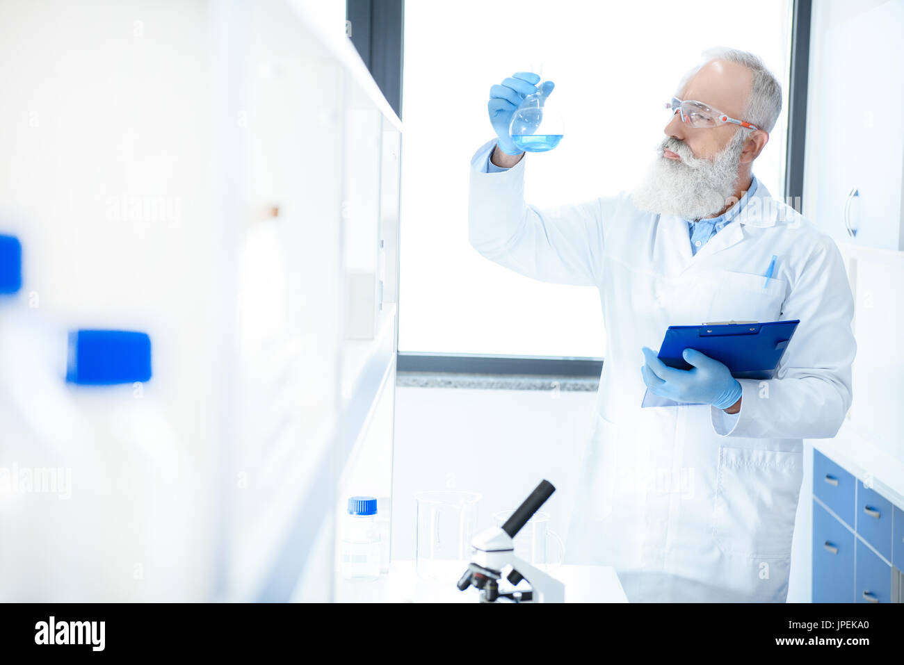 Senior bearded scientist holding flask with reagent and clipboard in ...