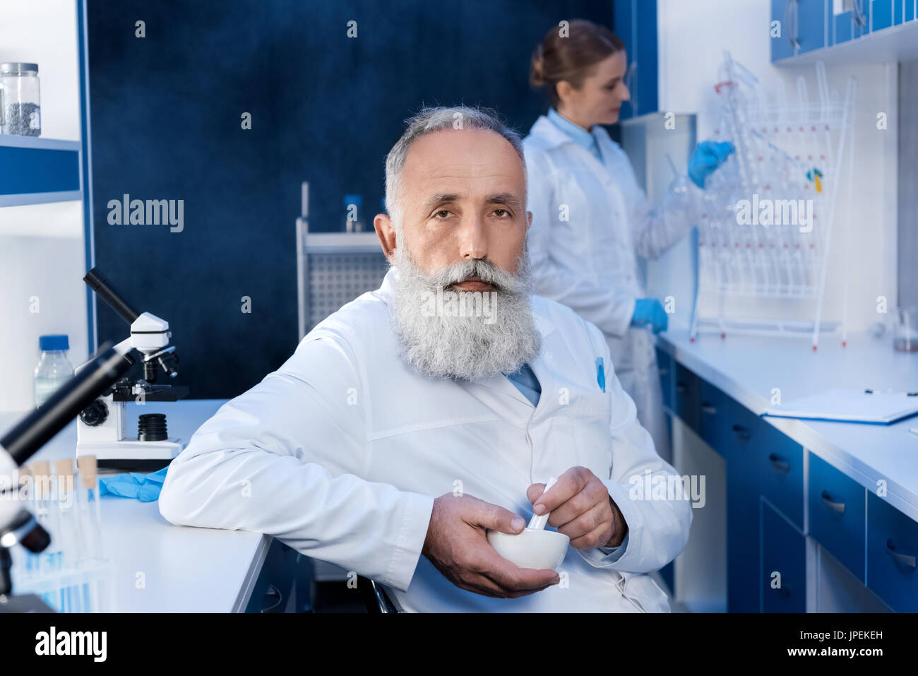 grey haired scientist sitting and looking at camera while woman working ...