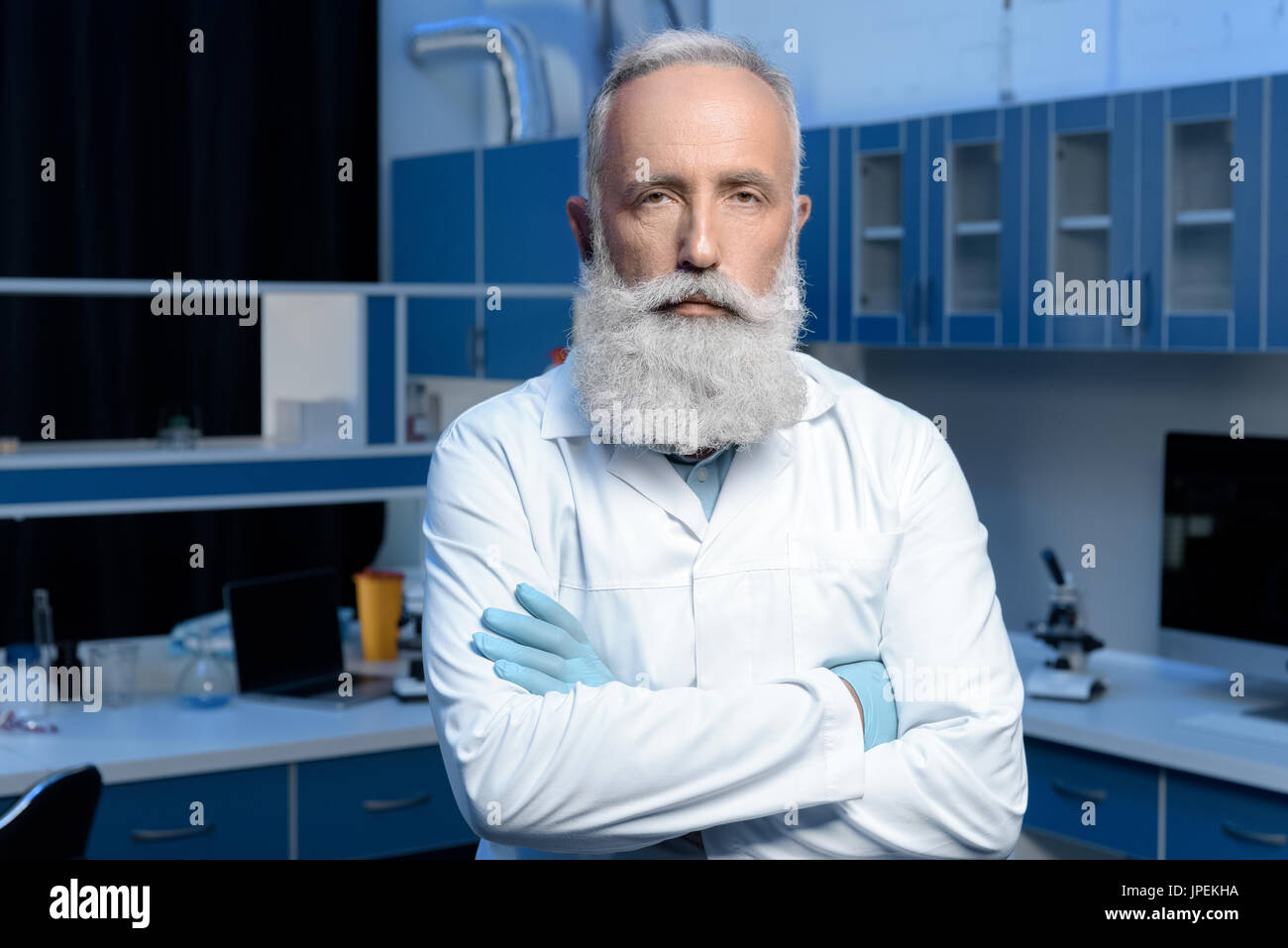 serious grey haired scientist in lab coat looking at camera with arms ...