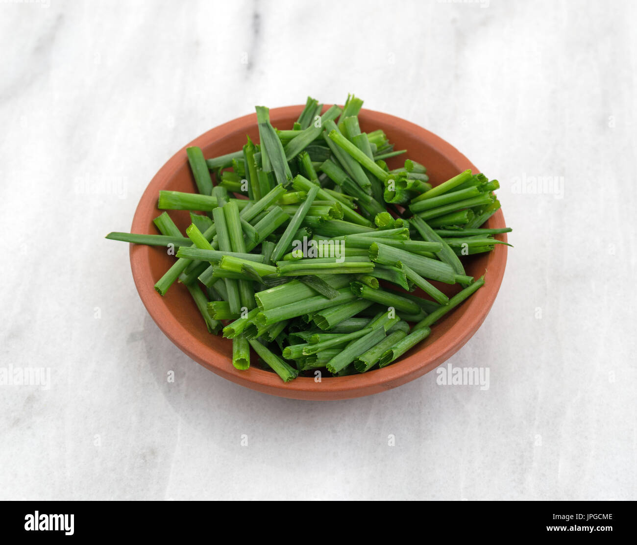 Cut organic chives in a small red clay bowl atop a gray marble cutting ...