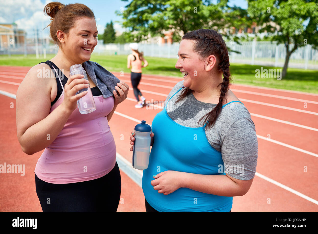 Laughing fat friendly women in sportswear drinking water on stadium ...