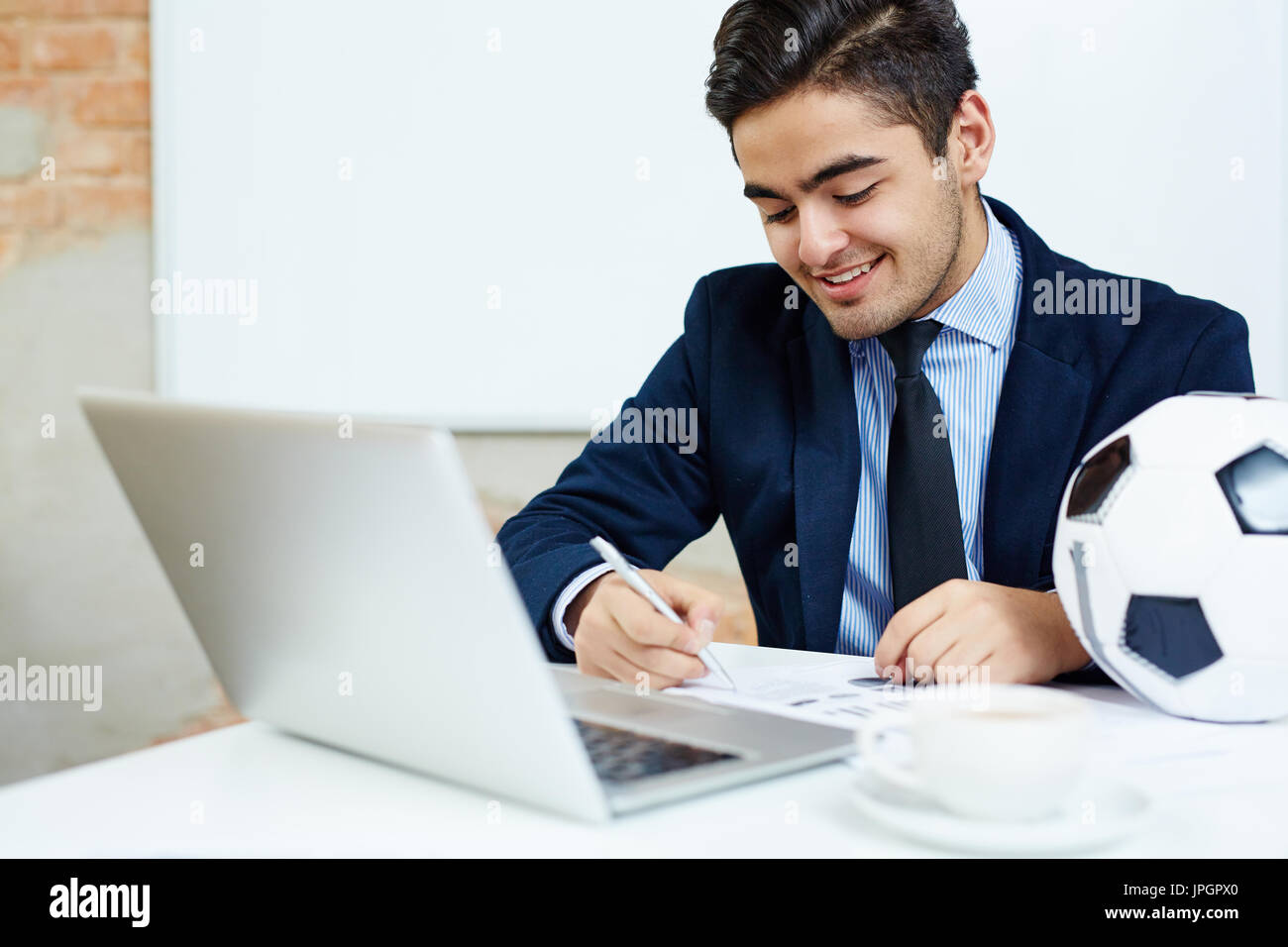 Young businessman making notes in front of laptop while waiting for ...