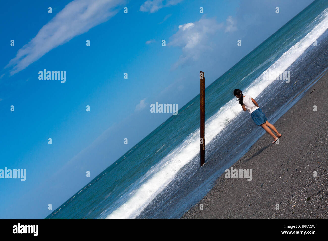 Girl standing facing inclined tide-pole on the beach by the sea ...