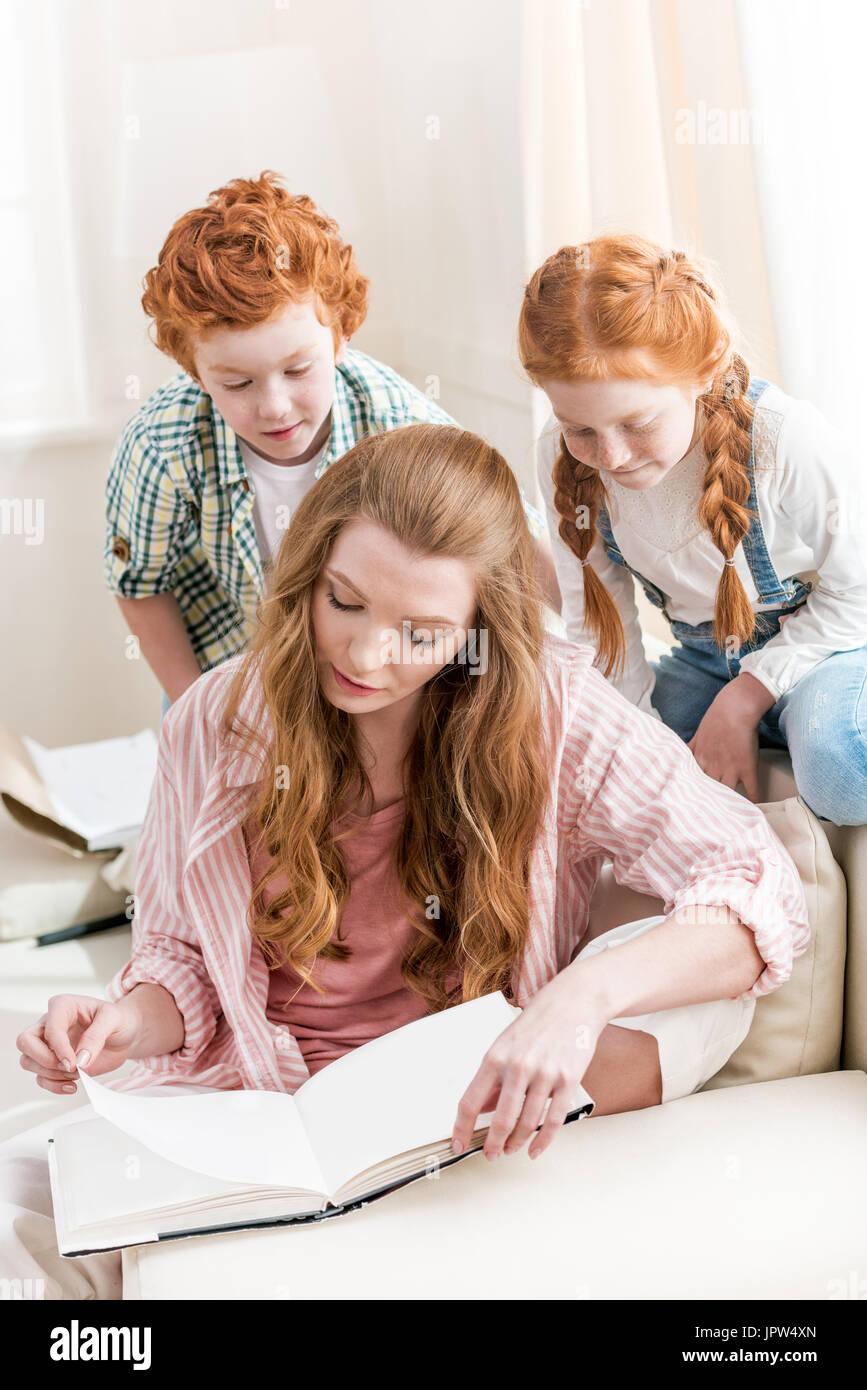 Happy mother with adorable redhead kids reading book together at home ...