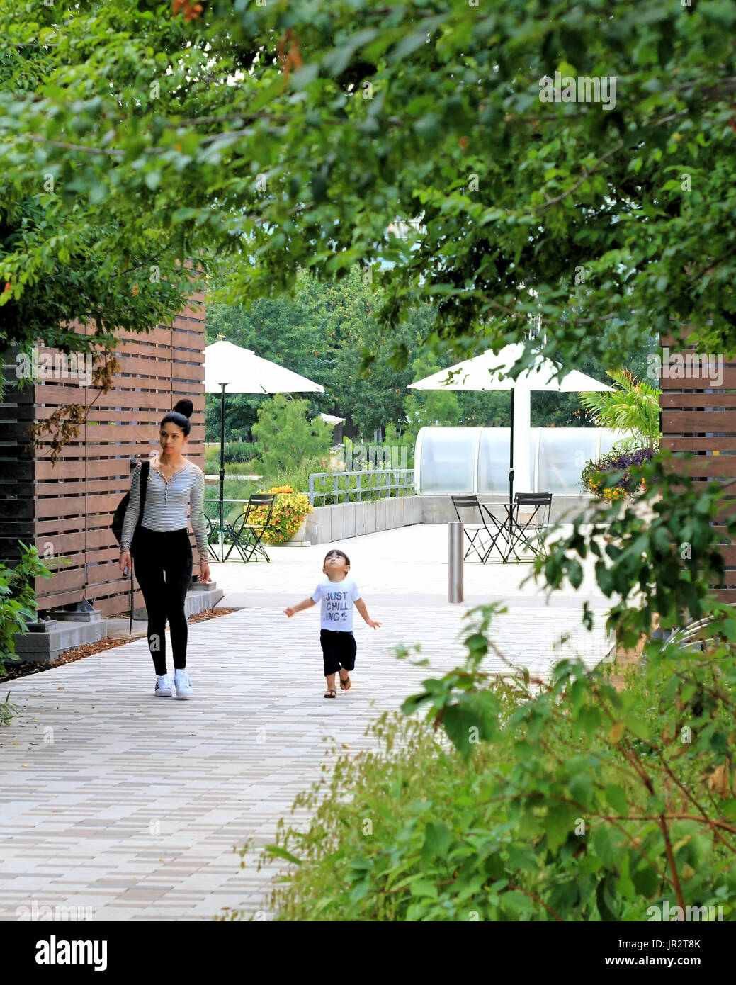 A young woman and a toddler boy walk on sidewalk near a garden cafe ...