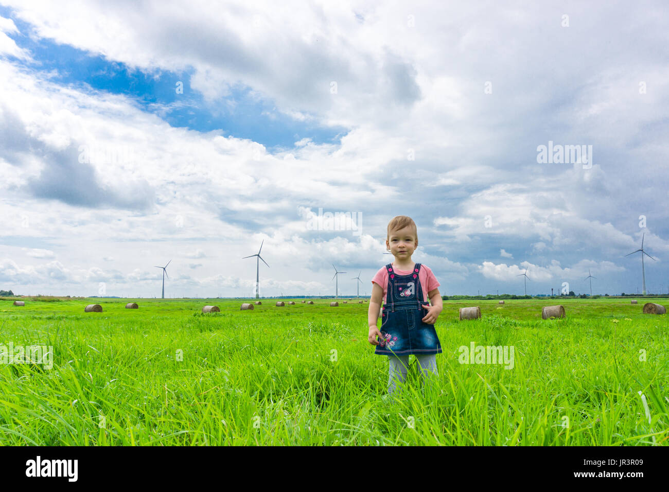little girl child is standing in green field with hay rolls in front of ...