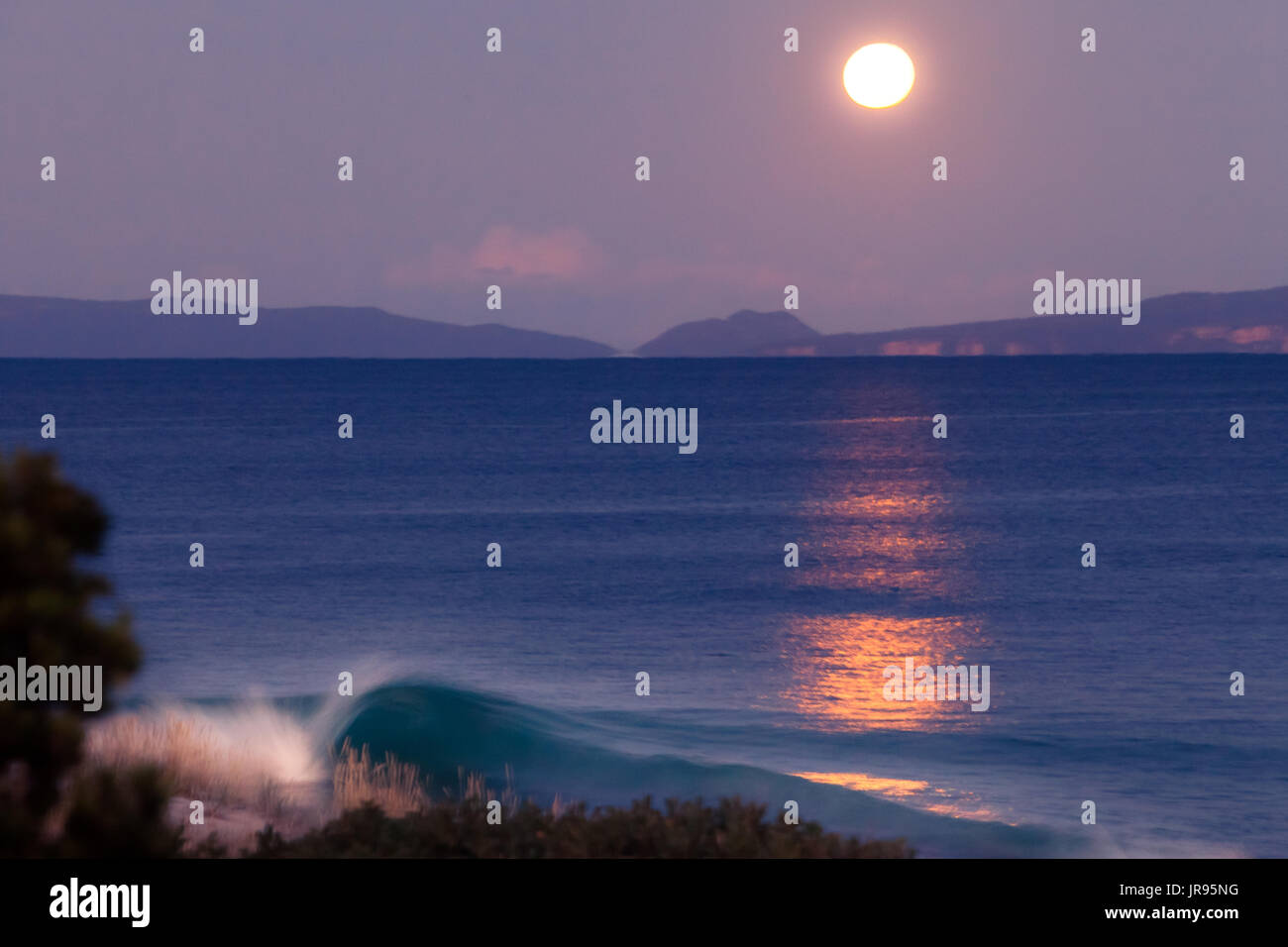 Full moon above the ocean with a crashing wave and the moon lighting up ...