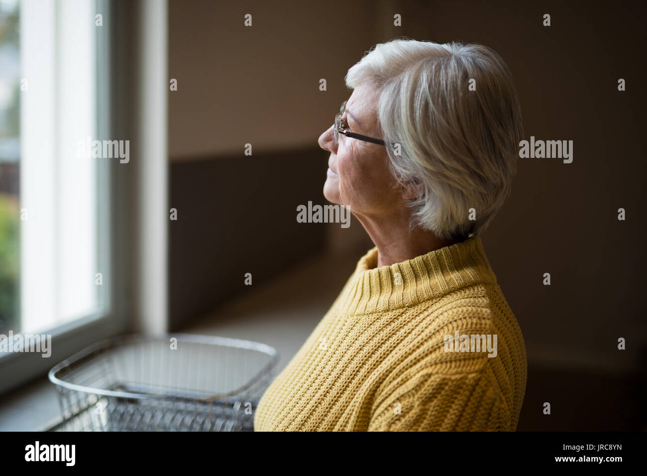 Close-up of thoughtful senior woman looking through kitchen window ...