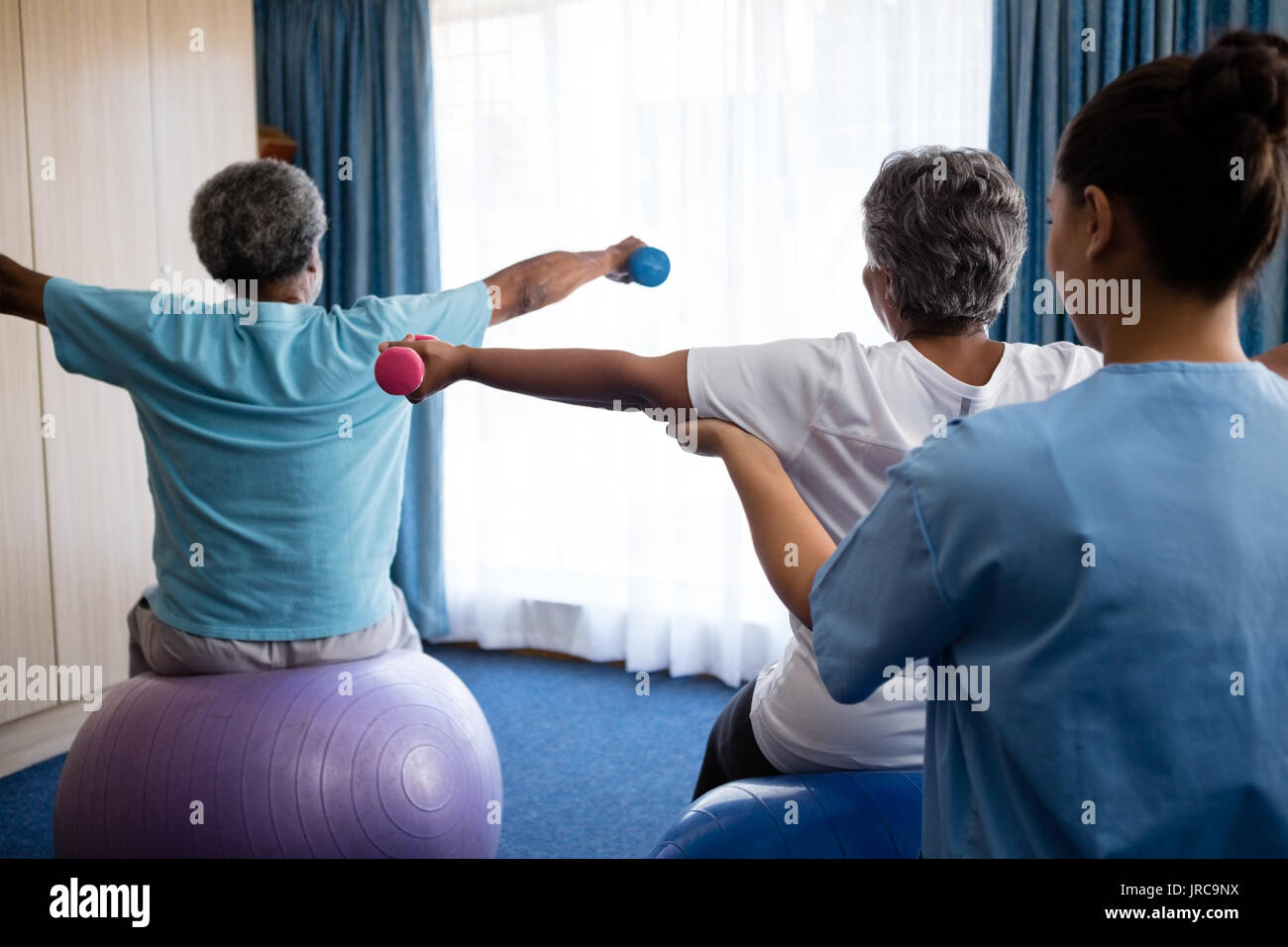 Rear view of nurse training seniors in lifting dumbbells while sitting ...