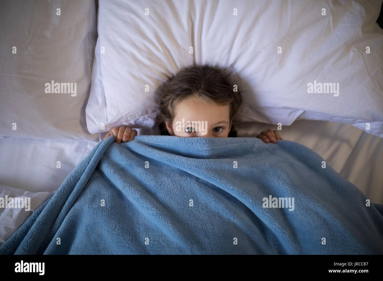 Portrait of girl covering her face under the blanket while lying on bed ...