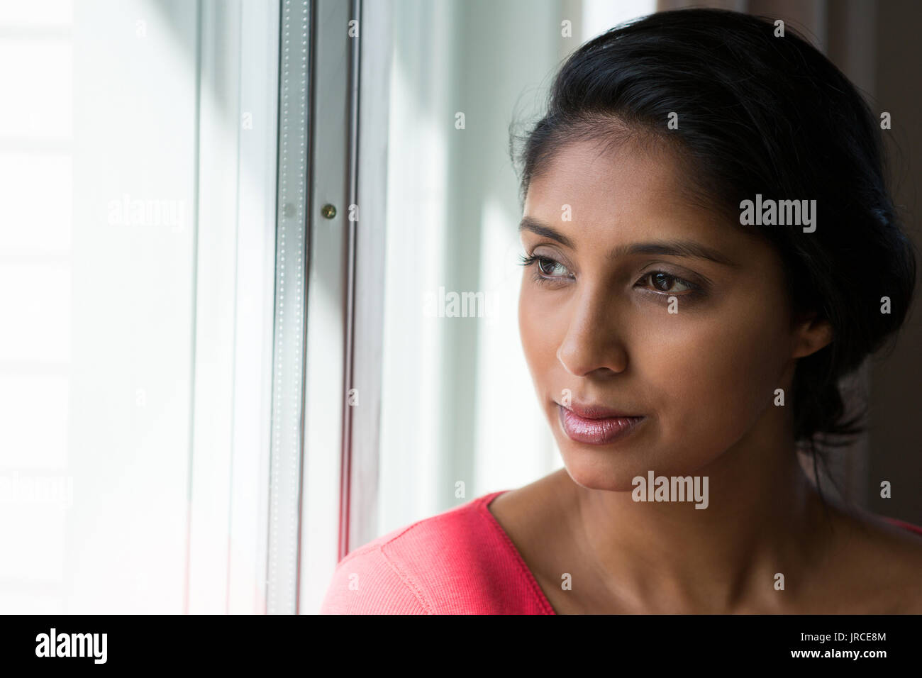 Young woman looking through window at home Stock Photo - Alamy