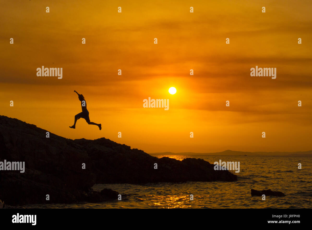 Man jumping rocky outcrop over sea at sunset in silhouette Stock Photo ...