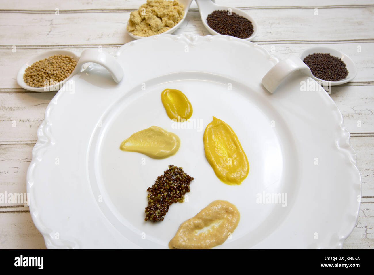 Smears of various mustards on white china plate with whole seeds and ...