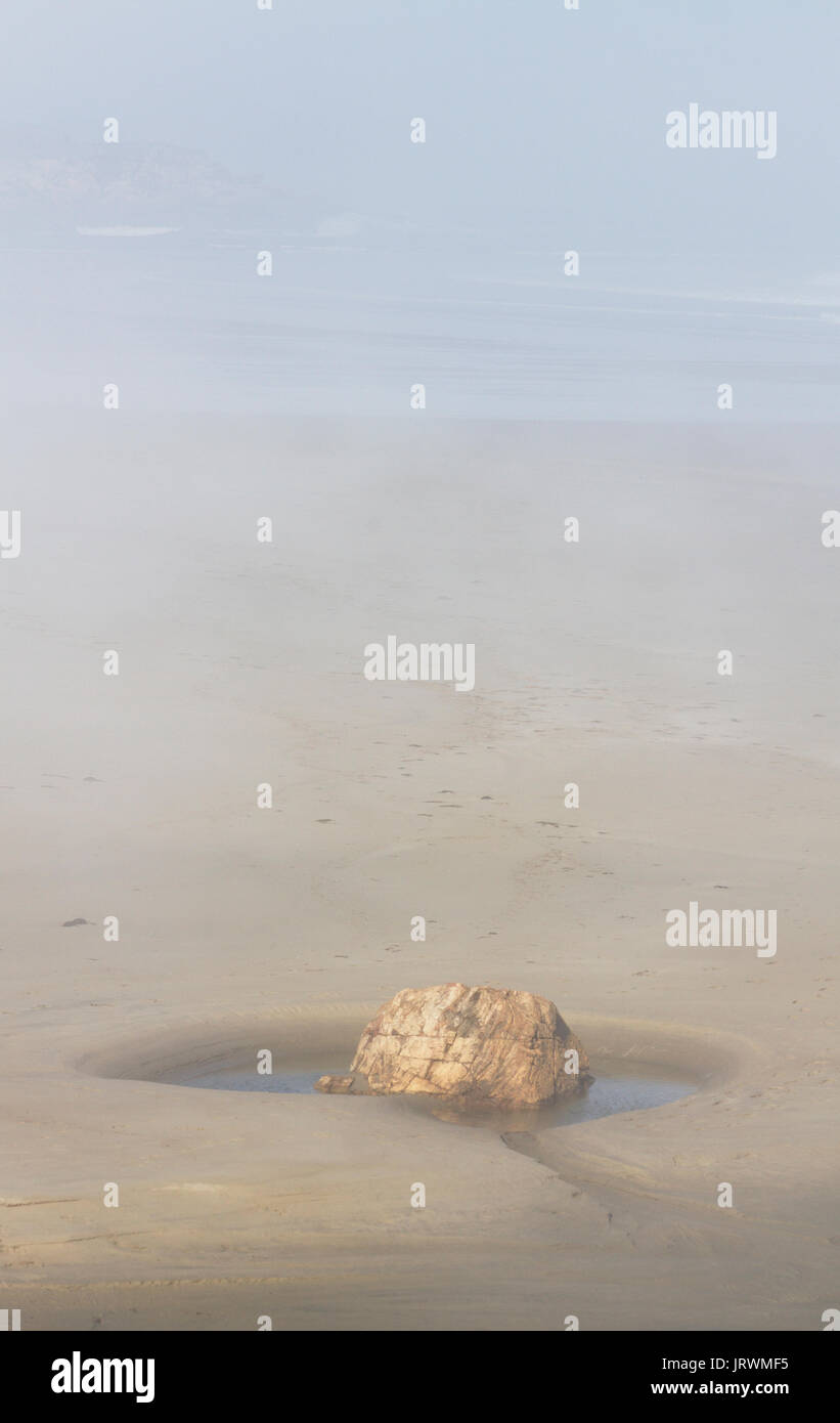 Large rock on the beach surrounded by water during high tide on a foggy ...